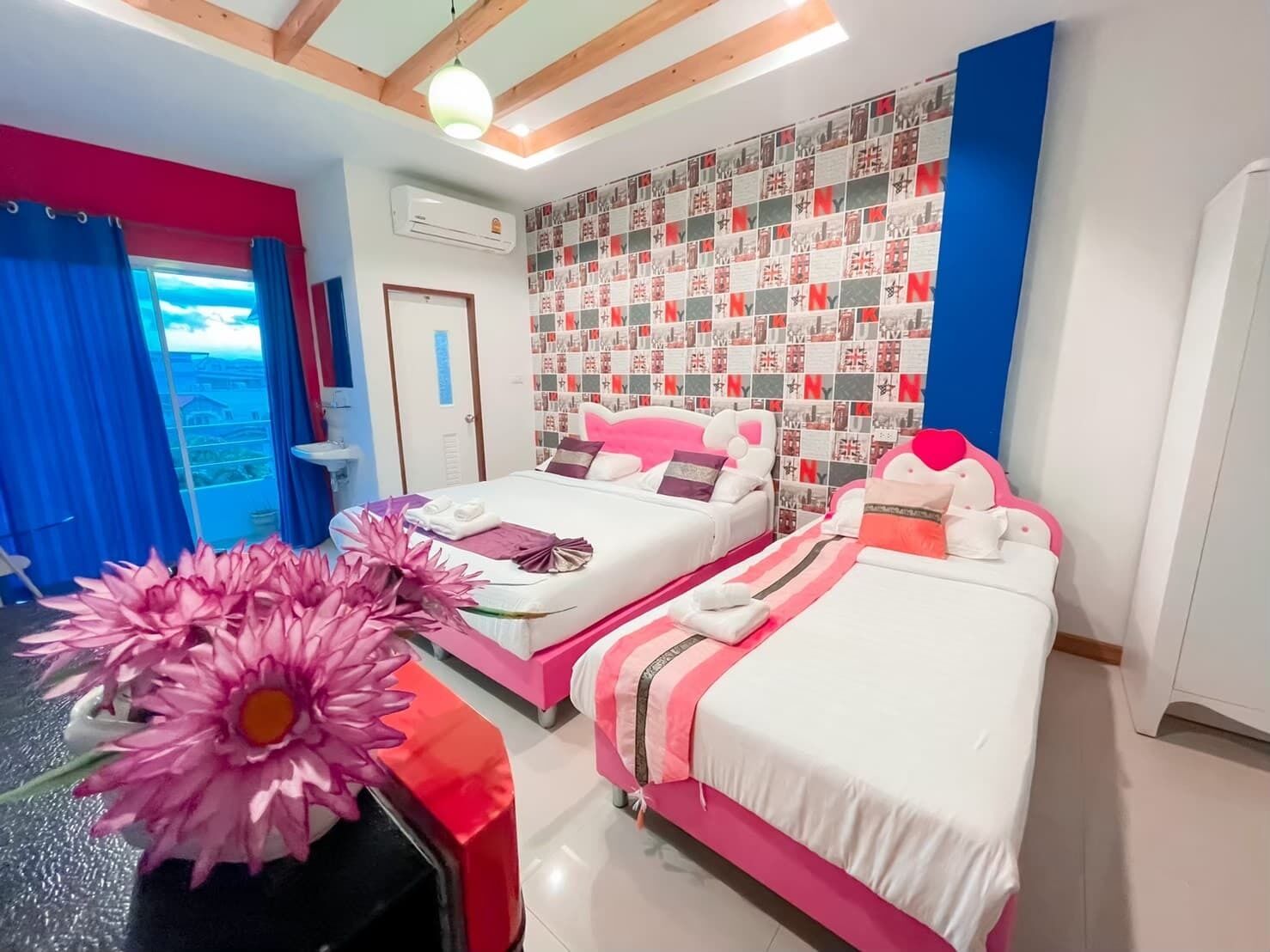 Quarto de hotel com duas camas, uma janela grande com vista, parede decorada com colcha de retalhos e um vaso de flores no chão.