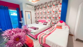 Quarto de hotel com duas camas, uma janela grande com vista, parede decorada com colcha de retalhos e um vaso de flores no chão.