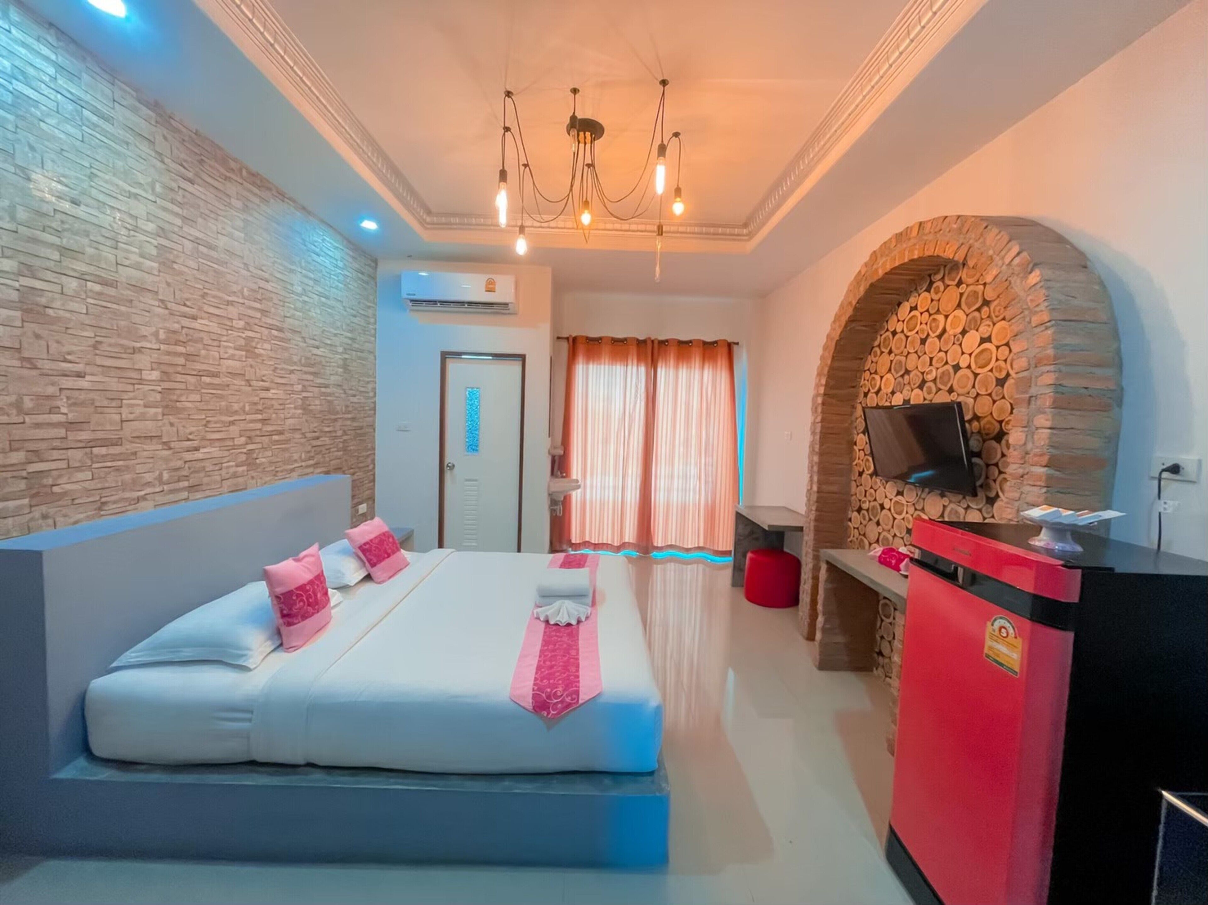 Quarto de hotel com uma cama grande, uma escrivaninha com um armário vermelho, uma TV fixada numa parede de pedra e um candelabro.