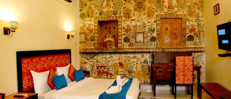Quarto de hotel com uma cama grande, uma escrivaninha e uma cadeira. Uma parede exibe uma obra de arte detalhada e há uma televisão na parede.