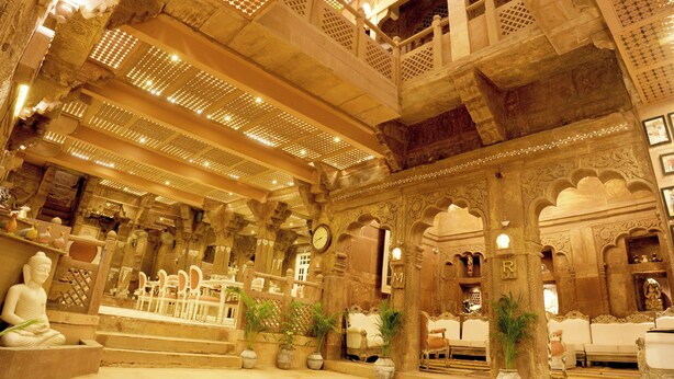 Interior ornamentado com colunas de pedra, uma estátua de Buda e um espaço de estar com cadeiras e mesas.