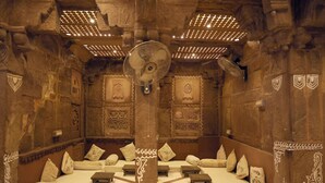 Quarto com paredes de pedra, um pilar central e várias áreas de estar com almofadas.