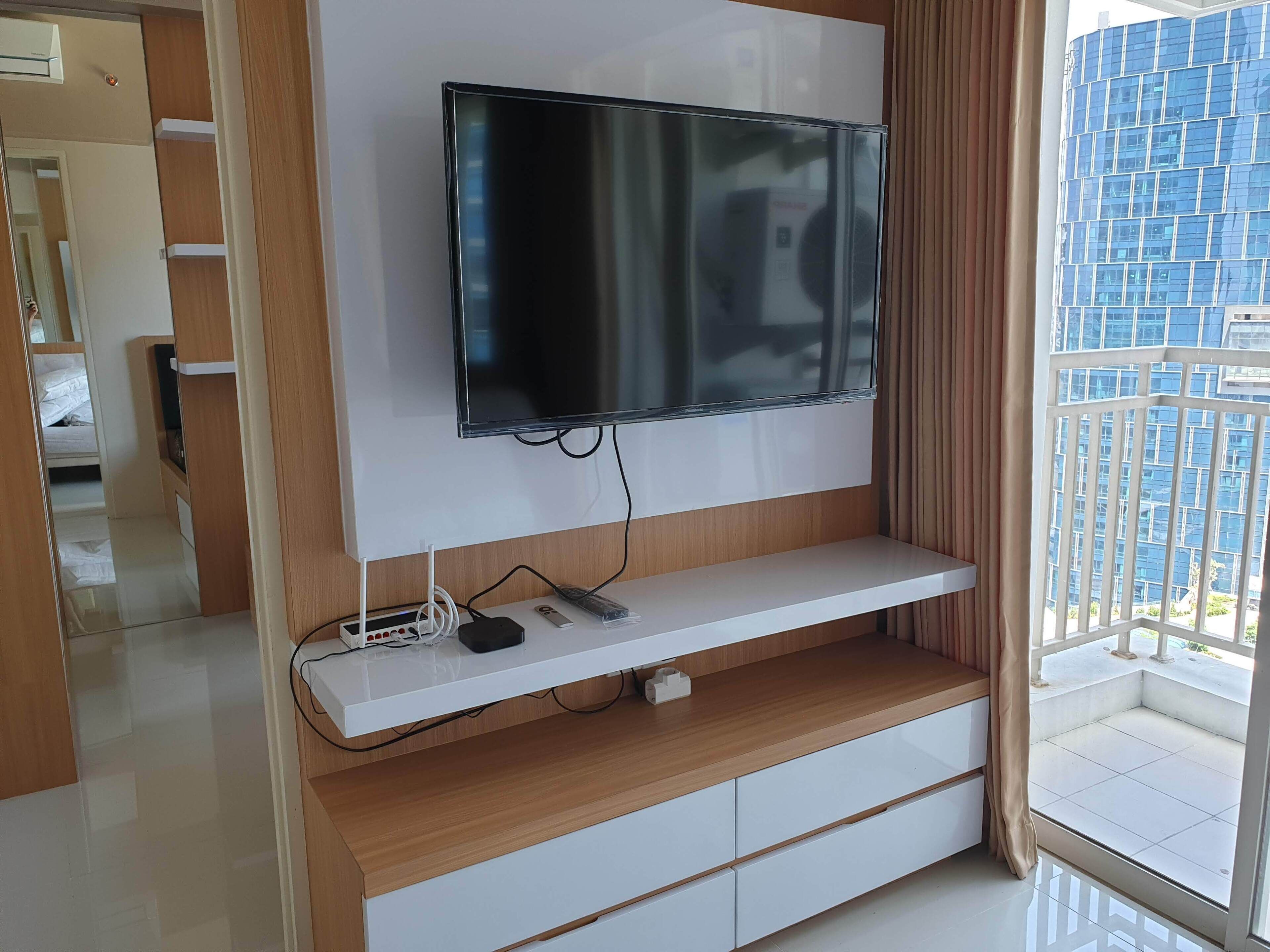 Uma sala de estar moderna com uma TV de tela plana fixada em uma parede de madeira, um console branco abaixo e uma varanda com vista para um edifício azul.