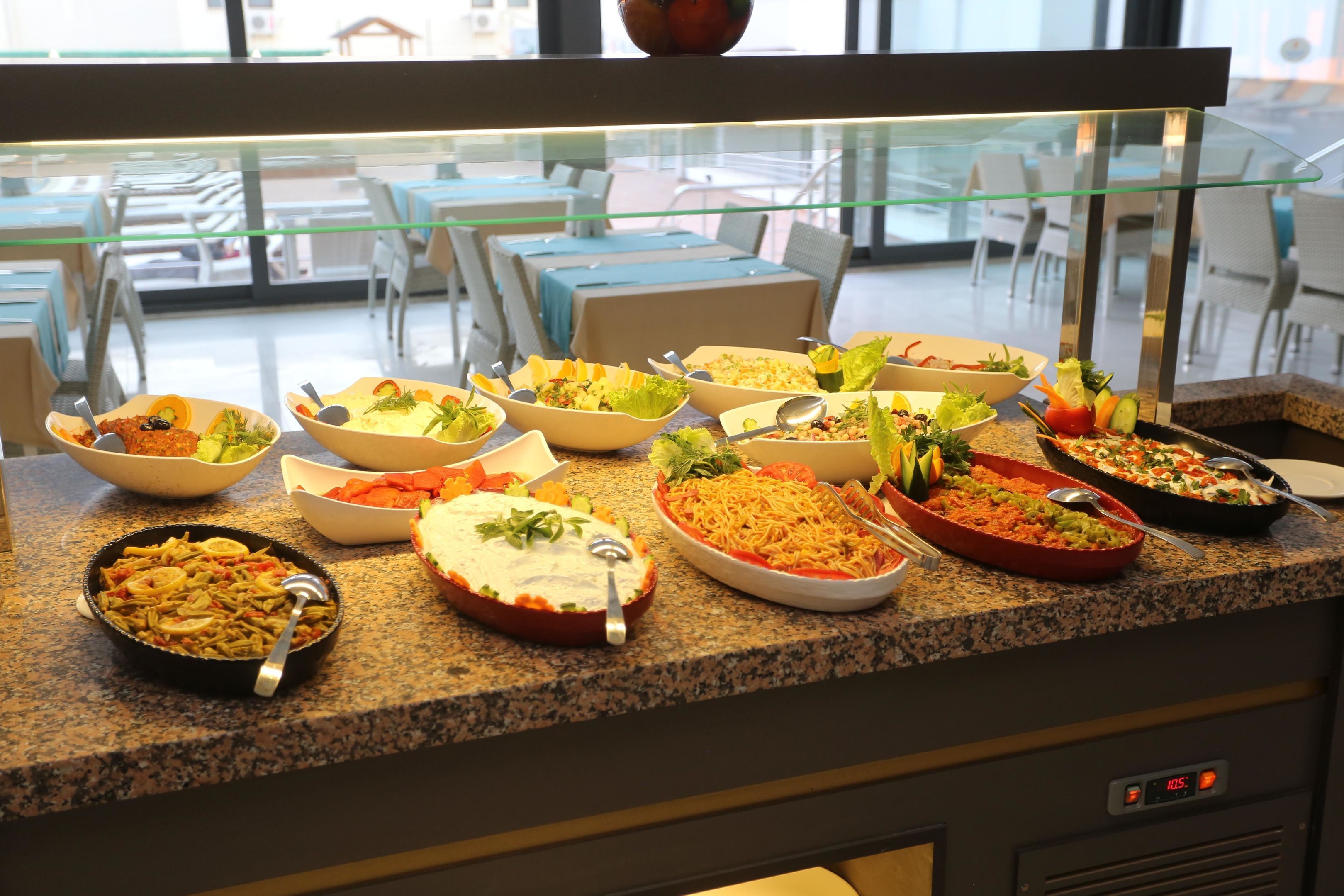 Buffet variado com diversos pratos, incluindo saladas, massas e um ensopado, com um balcão de vidro separando a comida da área de refeições.