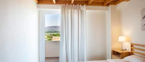 Quarto com teto de madeira, cama com roupa de cama branca, mesa de cabeceira com luminária e vista para a paisagem através de uma porta.