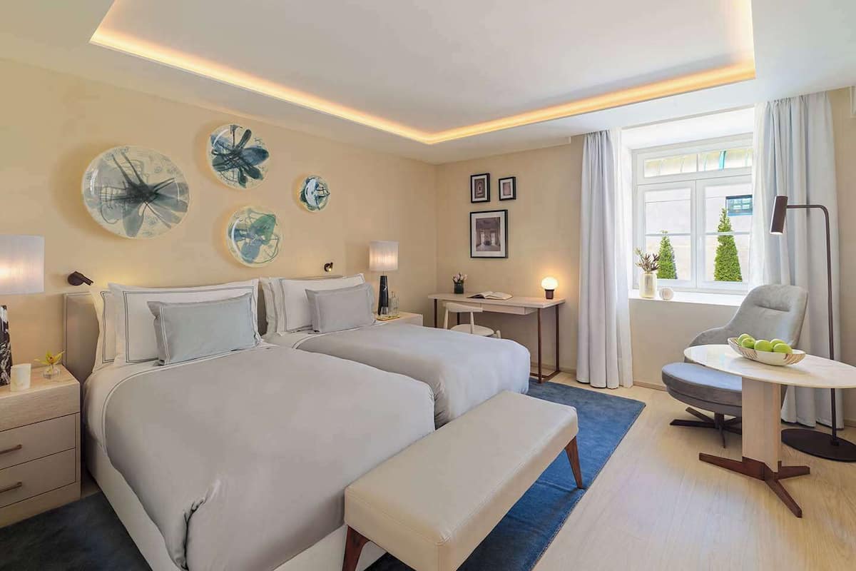 Quarto de hotel moderno com duas camas, uma escrivaninha e uma cadeira. Há pratos decorativos na parede e uma janela com cortinas.