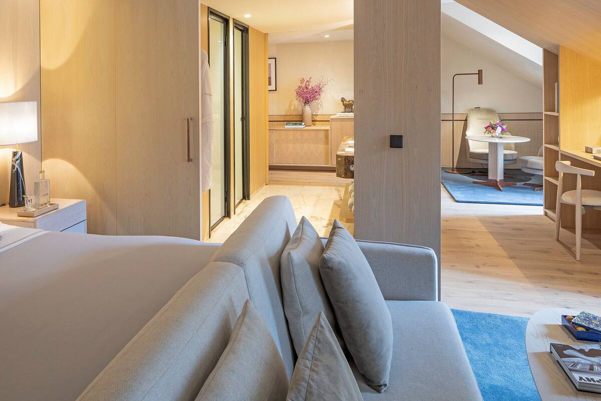 Quarto de hotel moderno com uma cama grande, área de estar com sofá e poltronas, uma pequena mesa de jantar, e vista para a cozinha e sala de estar.