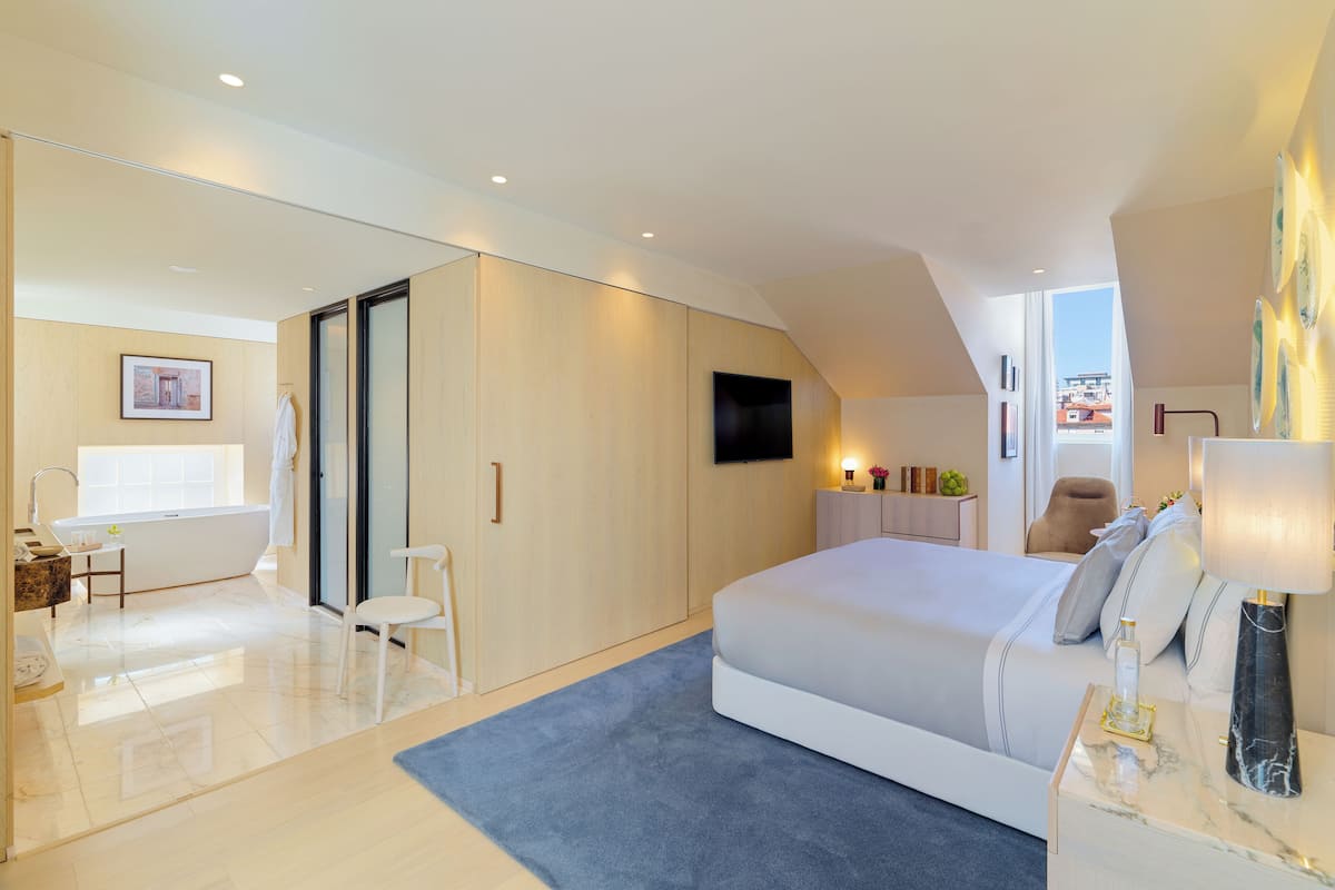 Quarto moderno com cama grande, área de estar com poltrona, banheira e televisão fixada na parede.