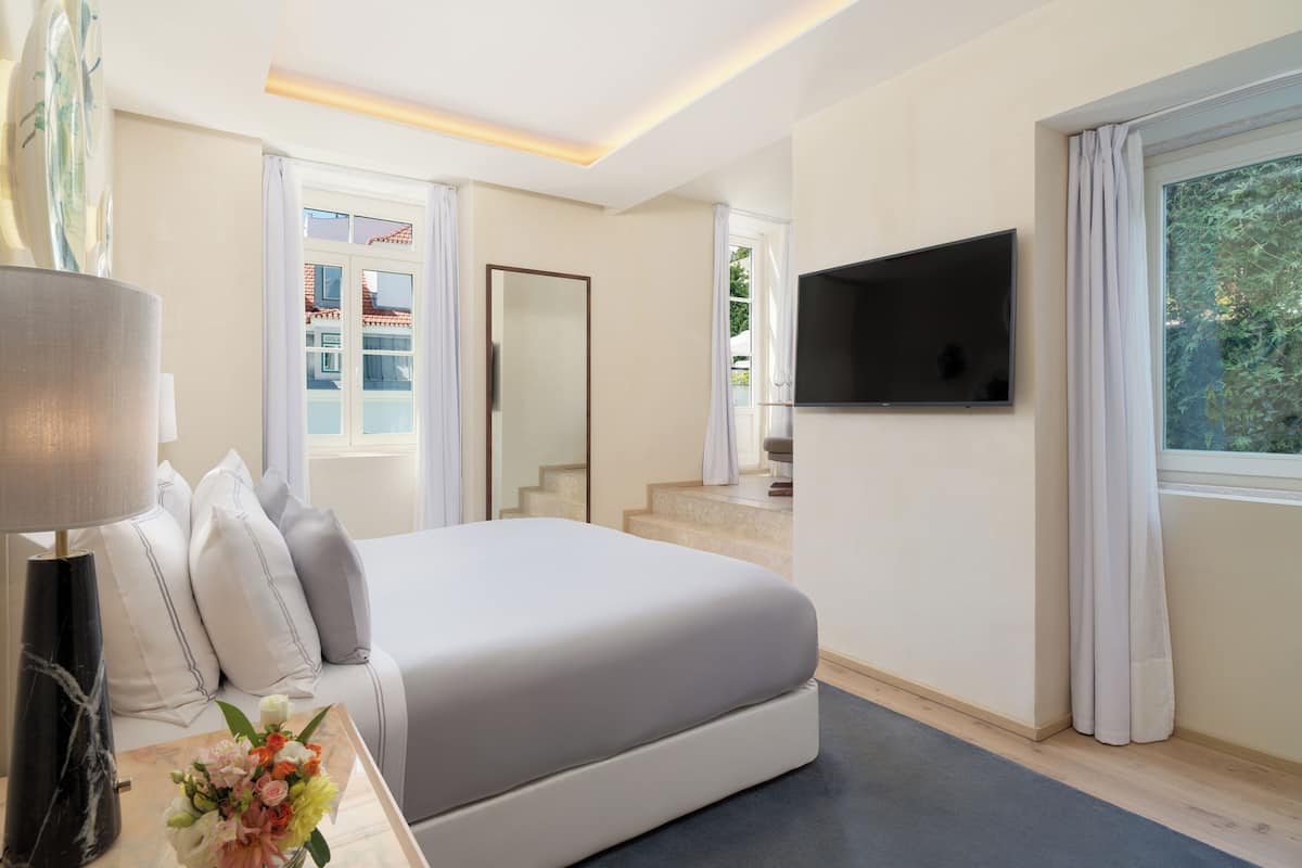 Quarto de hotel moderno com uma cama grande, televisão de tela plana, mesa de cabeceira com abajur e janela com cortinas.
