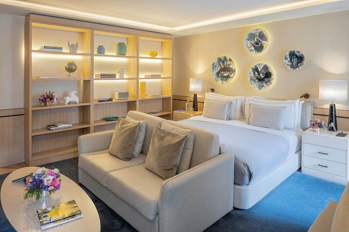 Quarto de hotel com cama, sofá, mesa de cabeceira, escrivaninha e estante.