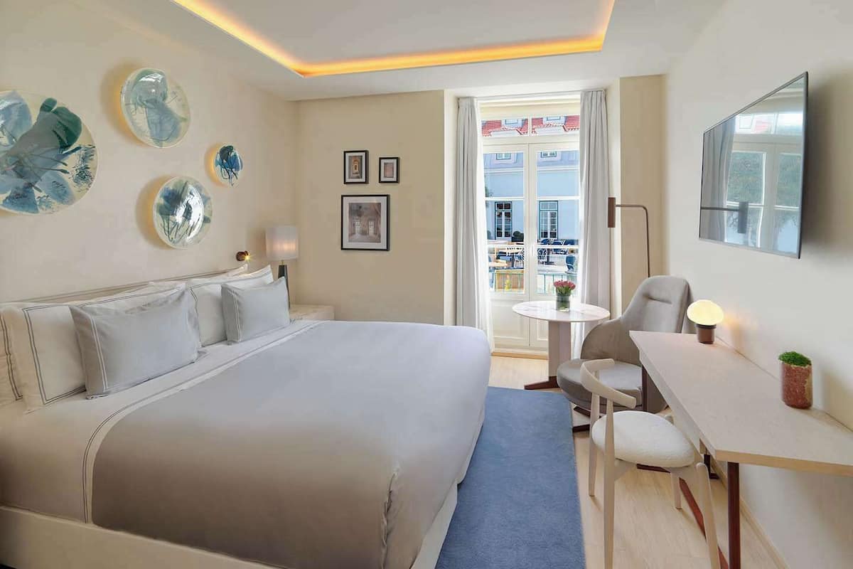 Quarto de hotel moderno com uma cama grande, uma escrivaninha e uma cadeira. Há pratos decorativos na parede, uma janela com cortinas e uma televisão de tela plana fixada na parede.