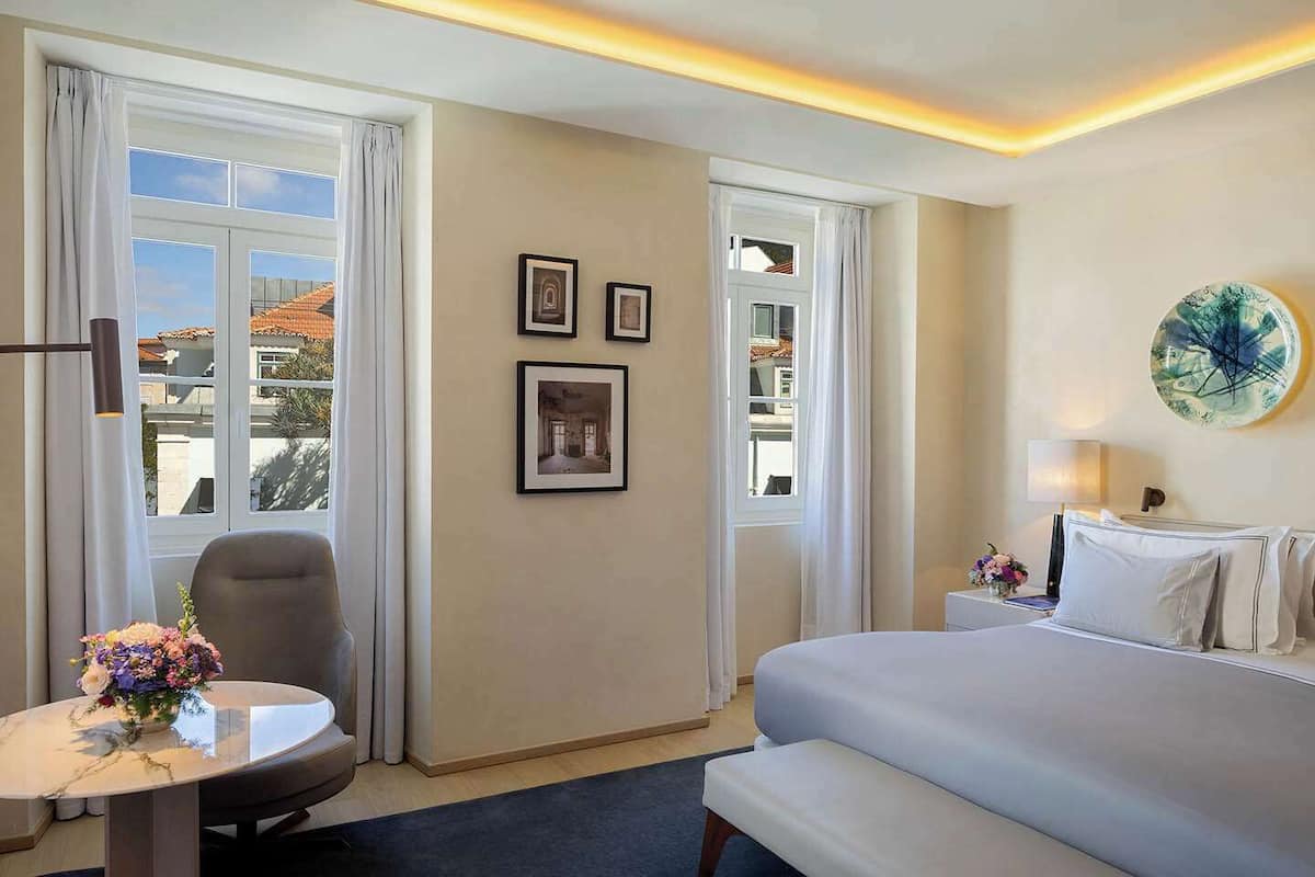 Quarto de hotel com cama, escrivaninha e cadeira, um vaso de flores e quadros nas paredes.