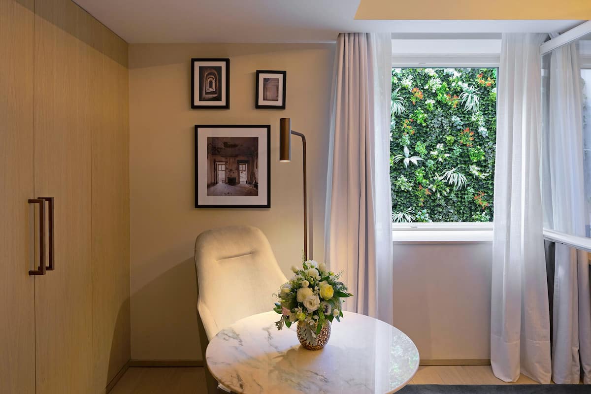 Um quarto com uma cadeira branca, uma mesa redonda com um vaso de flores, um quadro na parede e uma janela com cortinas.