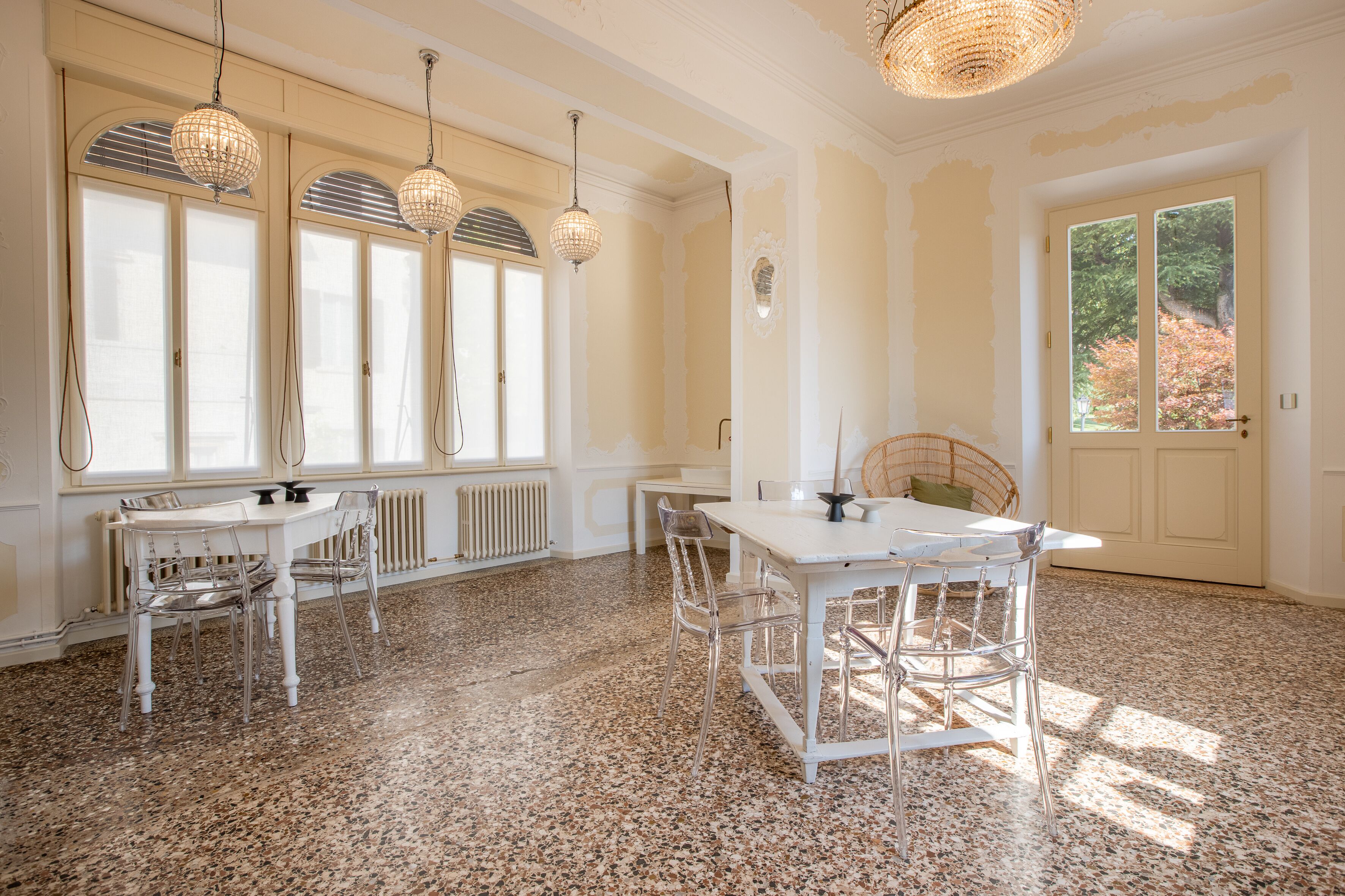 Um quarto espaçoso com uma grande mesa de jantar, cadeiras transparentes e um lustre. Há grandes janelas com persianas e uma porta que dá para o exterior.