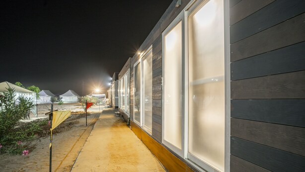 Uma fileira de cabanas com janelas amplas, um caminho de areia e um mastro.