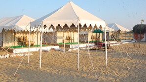 Uma série de tendas brancas com detalhes verdes numa praia arenosa, cada uma com uma pequena mesa e cadeiras.