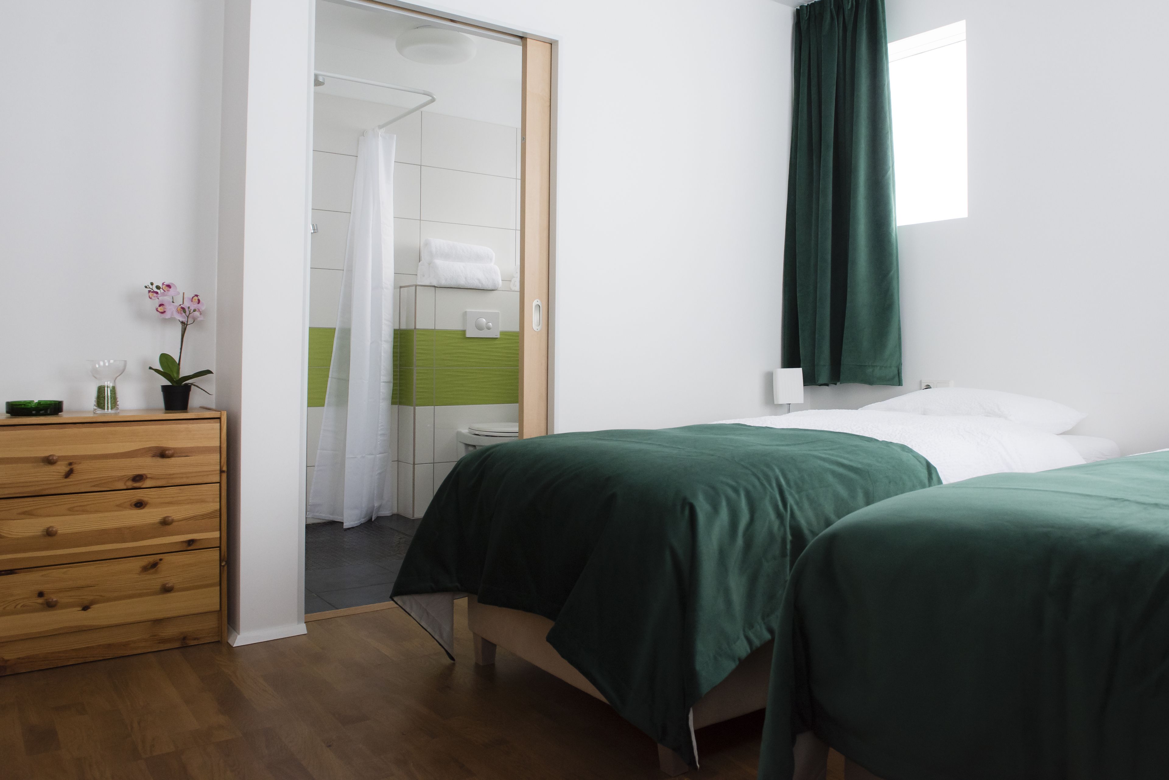Quarto com duas camas, cômoda de madeira, uma planta em vaso e banheiro visível por uma porta aberta.