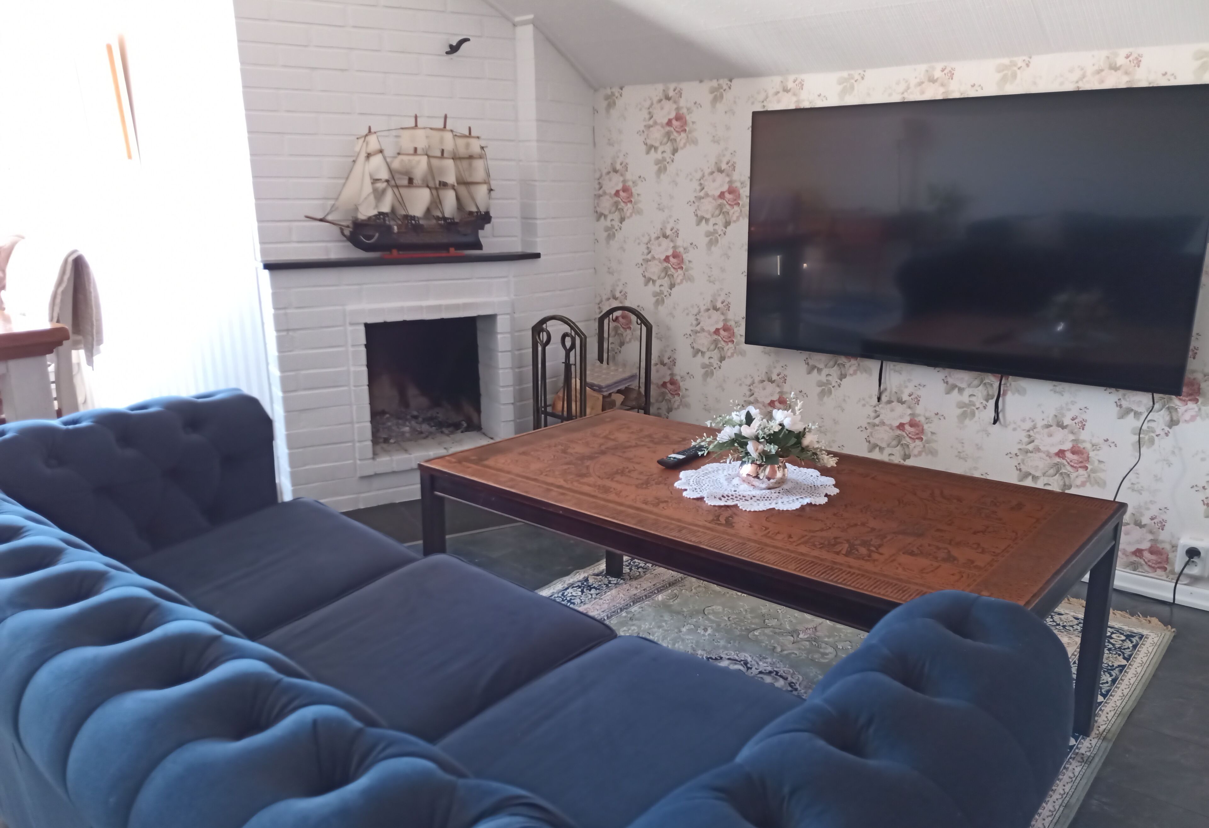 Uma sala de estar com um amplo sofá azul, uma mesa de centro de madeira com um vaso de flores, uma lareira com um modelo de navio e uma TV de tela plana fixada na parede.
