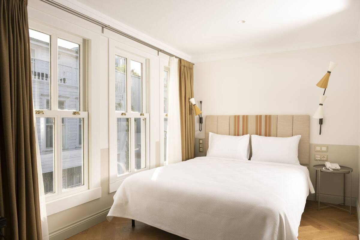 Quarto de hotel com uma cama grande, mesa de cabeceira com abajur e janela com cortinas.