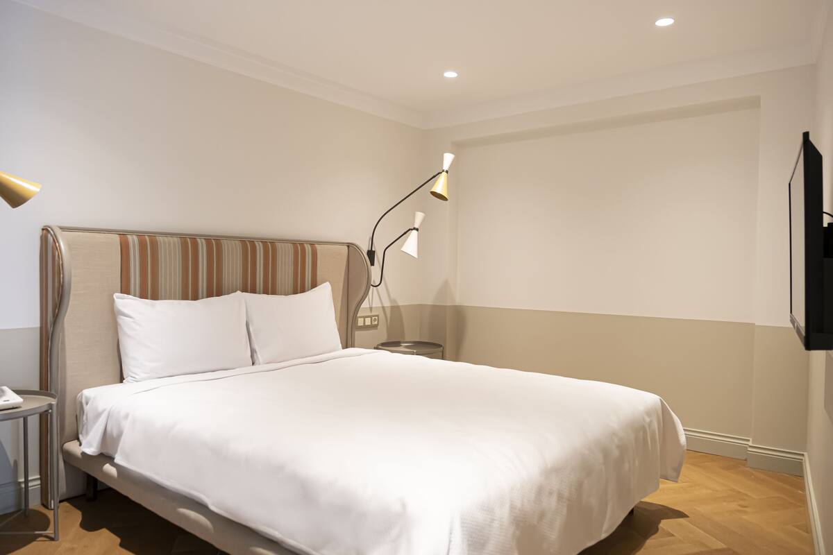Cama bem arrumada com lençóis brancos, mesa de cabeceira com abajur e televisão fixada na parede.