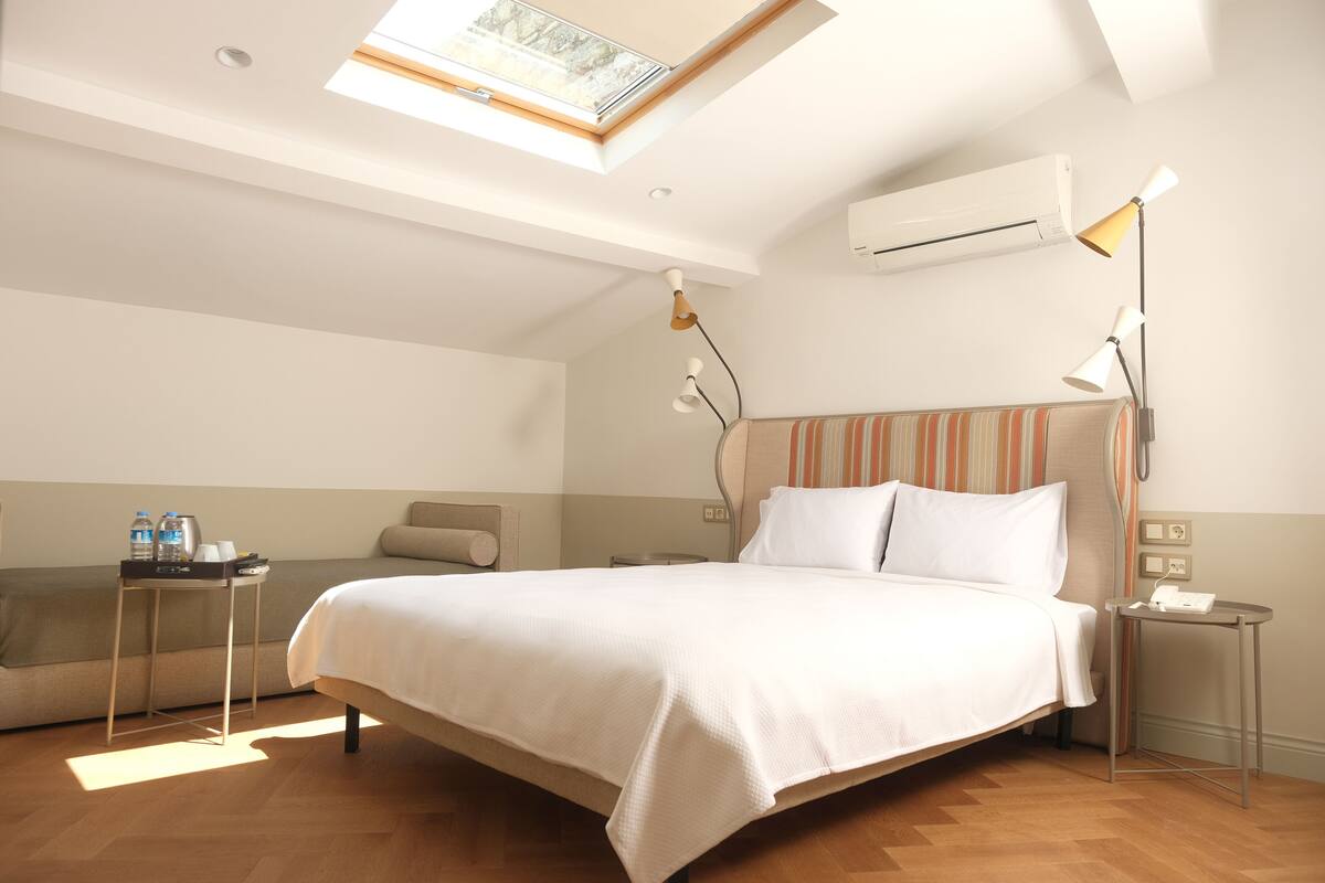 Quarto de hotel com cama, mesa de cabeceira com abajur, pequena mesa com garrafas de água e janela com vista para árvores.