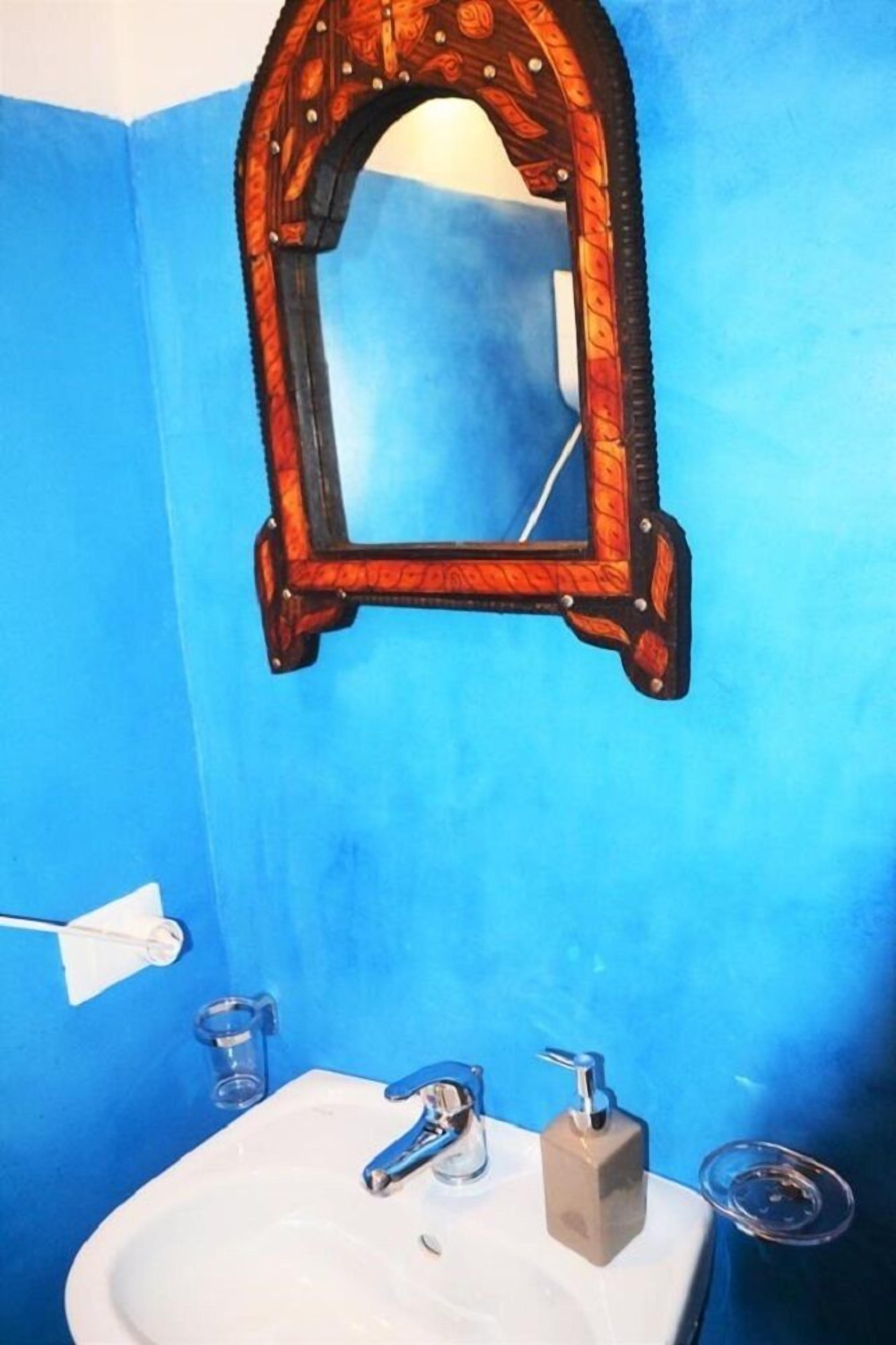 Uma casa de banho com parede azul, uma pia branca, um espelho com moldura de madeira e um dispensador de sabão.