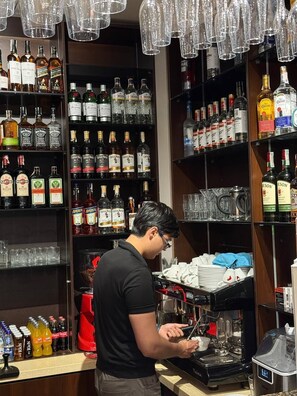 Uma área de bar com prateleiras repletas de garrafas de bebidas, uma máquina de café e copos pendurados.