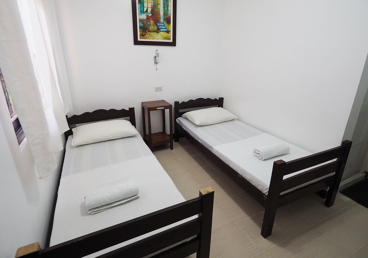 Quarto com duas camas de solteiro, uma pequena mesa de cabeceira e luminária fixada na parede.