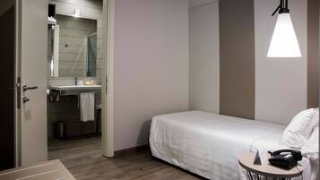 Quarto de hotel com uma cama, mesa de cabeceira com telefone, pequena mesa com um aviso, espelho e um banheiro visível através de uma porta aberta.