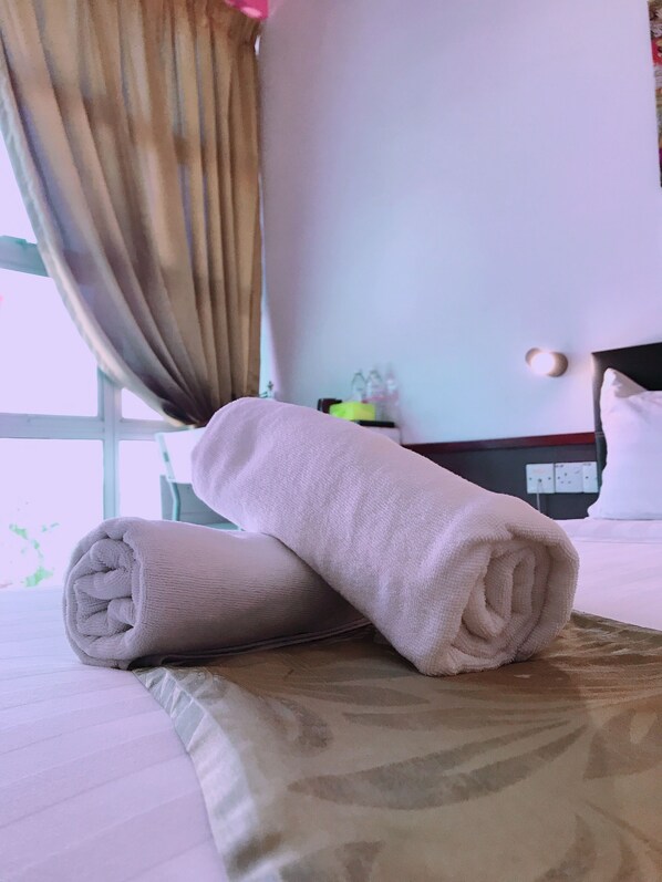 Uma cama bem arrumada com uma toalha enrolada, uma janela com cortinas e uma mesa de cabeceira com um abajur e uma garrafa.