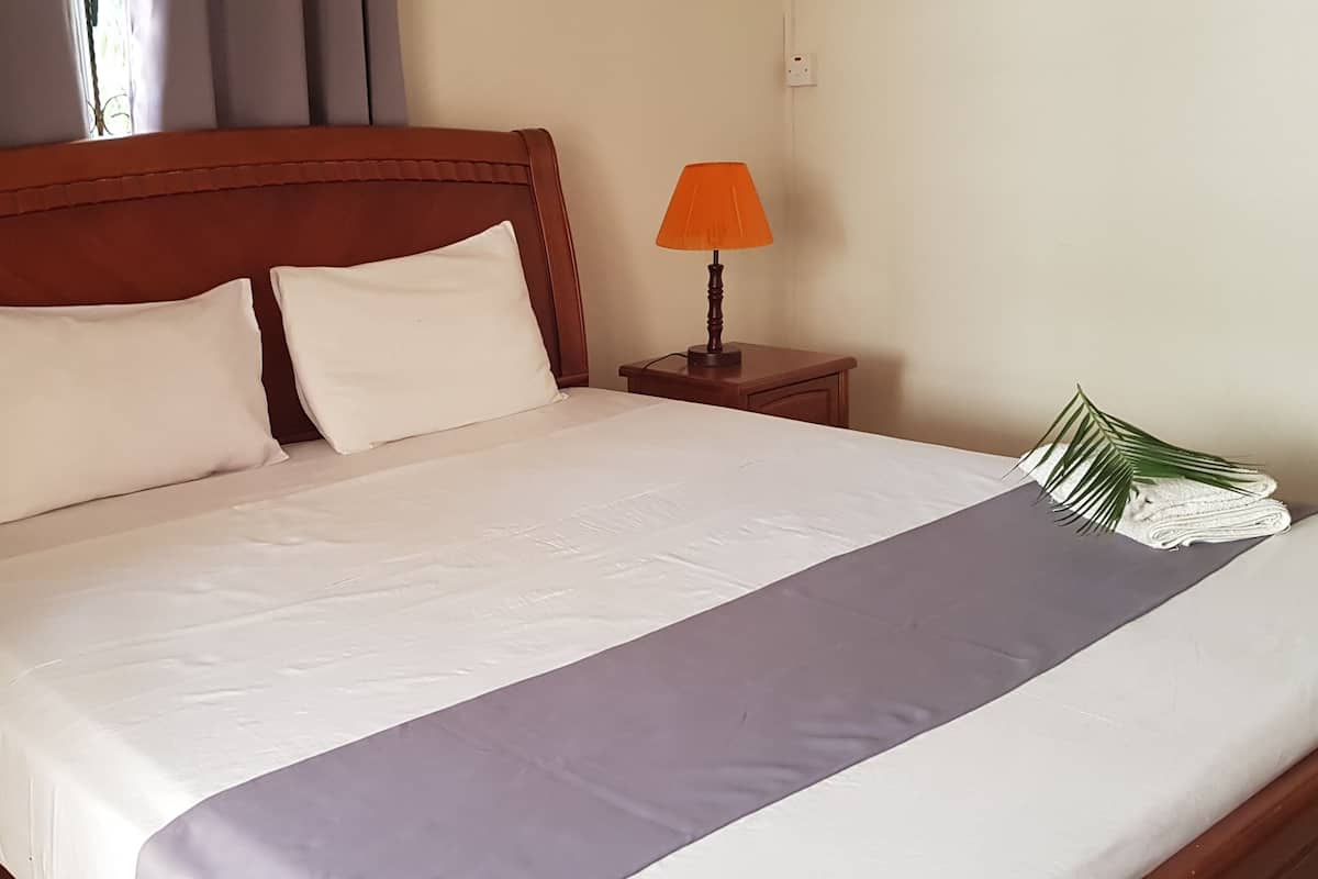 Cama bem arrumada com lençóis brancos e detalhes em roxo, mesa de cabeceira de madeira com abajur e toalha branca dobrada.