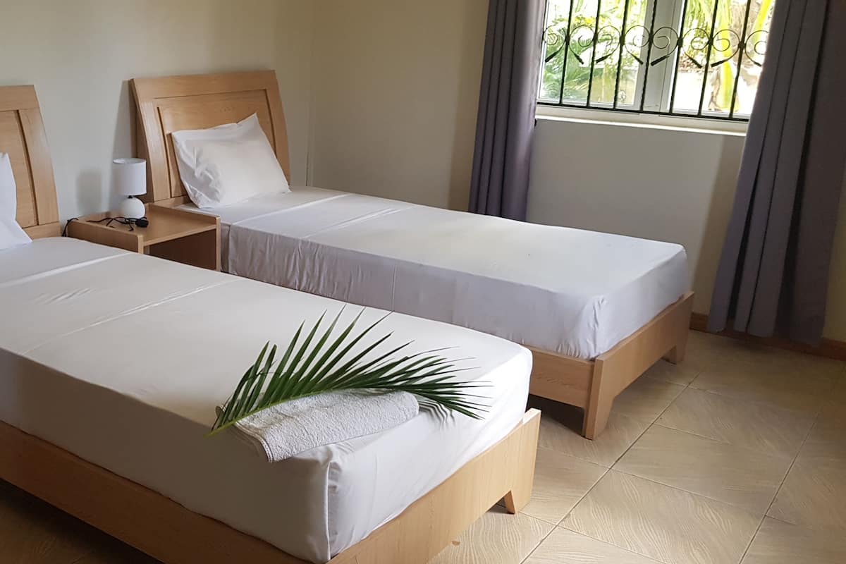 Duas camas de solteiro com cabeceiras de madeira, roupa de cama branca e uma pequena mesa de cabeceira com abajur.