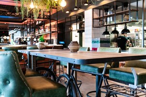 Interior moderno de restaurante com mesas altas, cadeiras estofadas verdes, plantas penduradas e uma área de bar ao fundo.