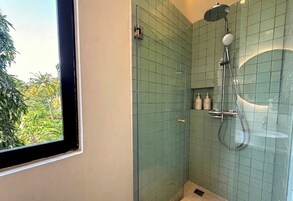 Uma casa de banho com azulejos verdes, chuveiro e janela com vista para a vegetação.