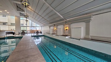 Piscina interior com água cristalina, rodeada por paredes azulejadas brancas e teto com padrão quadriculado.
