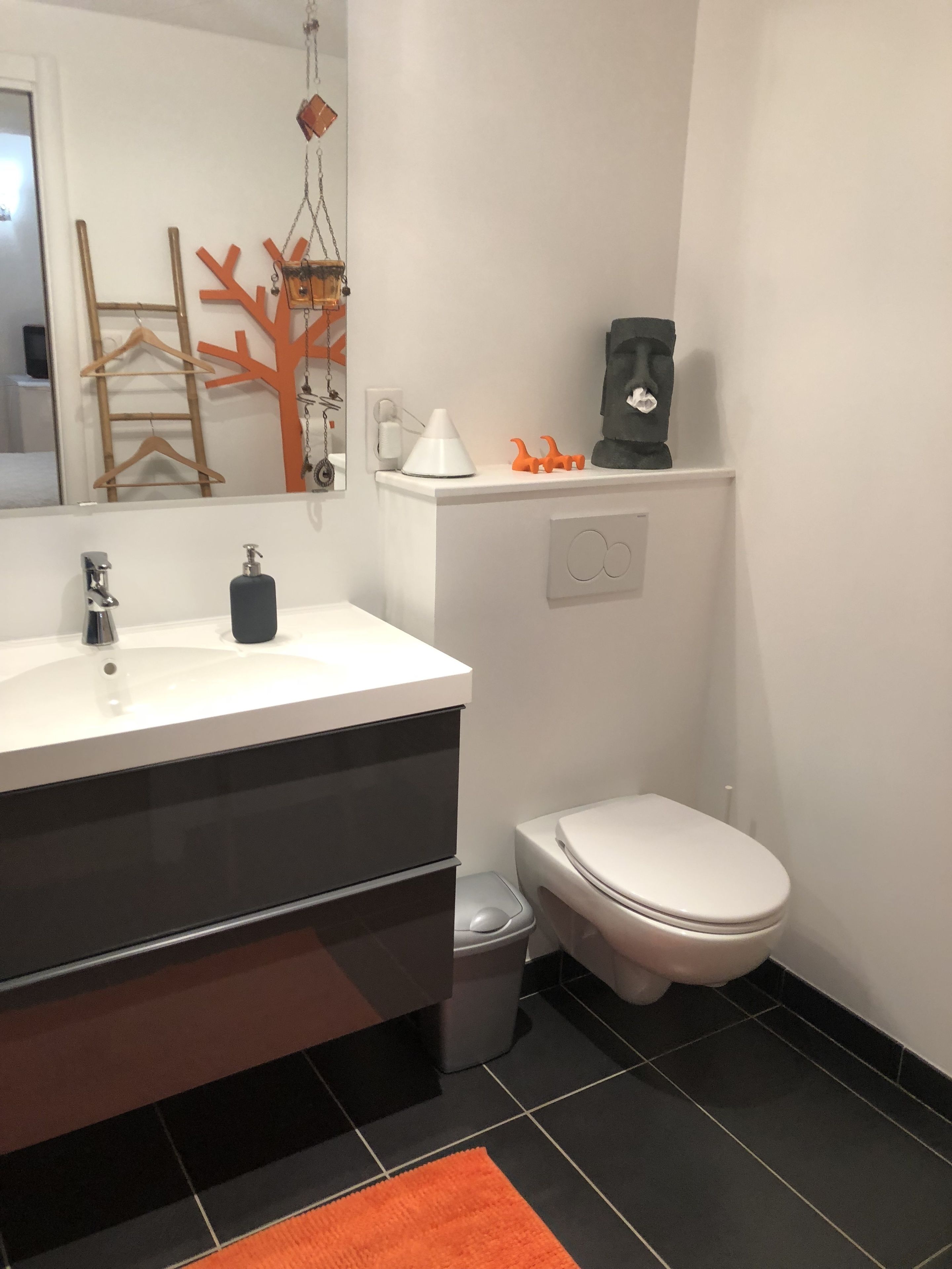 Casa de banho moderna com lavatório branco, armário preto e sanita branca. Há uma pequena prateleira com objetos decorativos e um candeeiro pendente sobre o lavatório.
