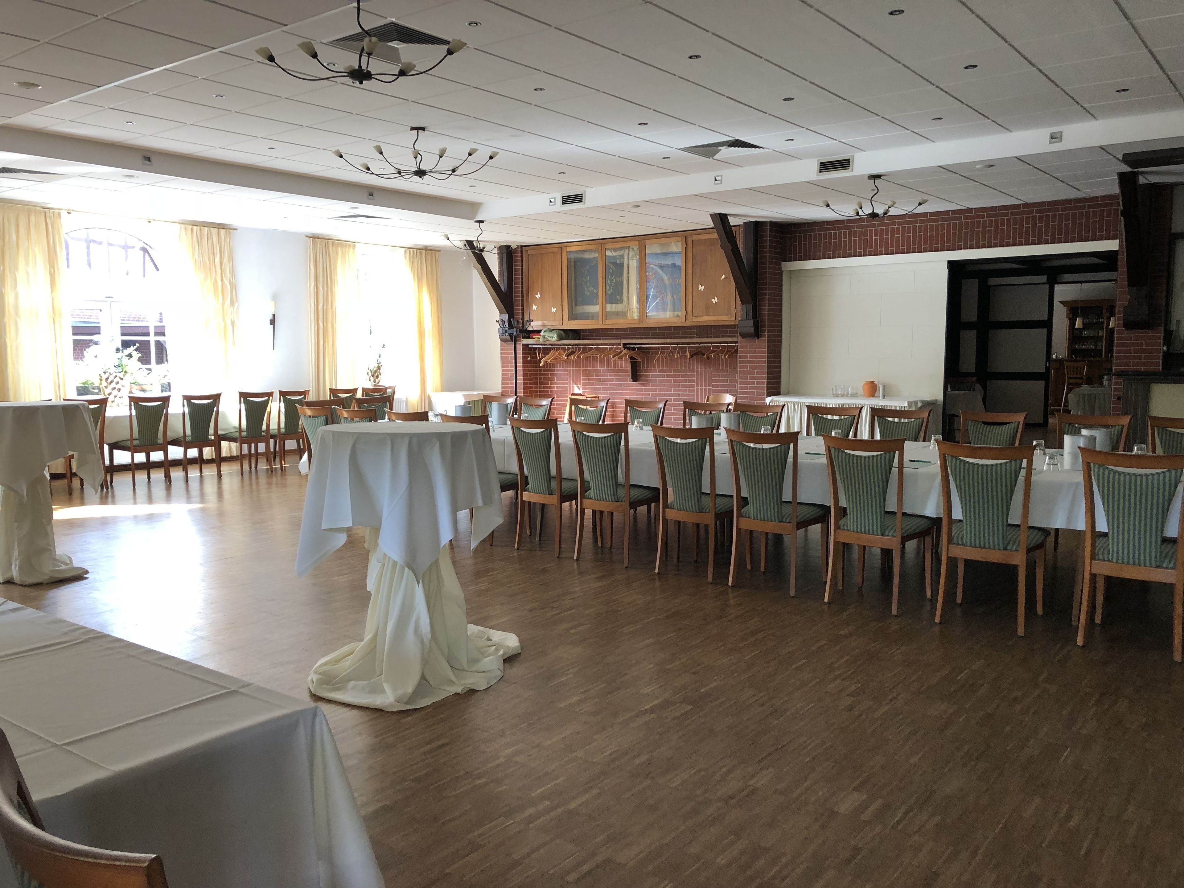 Uma sala de jantar ampla, com várias mesas e cadeiras, soalho de madeira e uma parede de tijolos, com uma área de cozinha visível ao fundo.