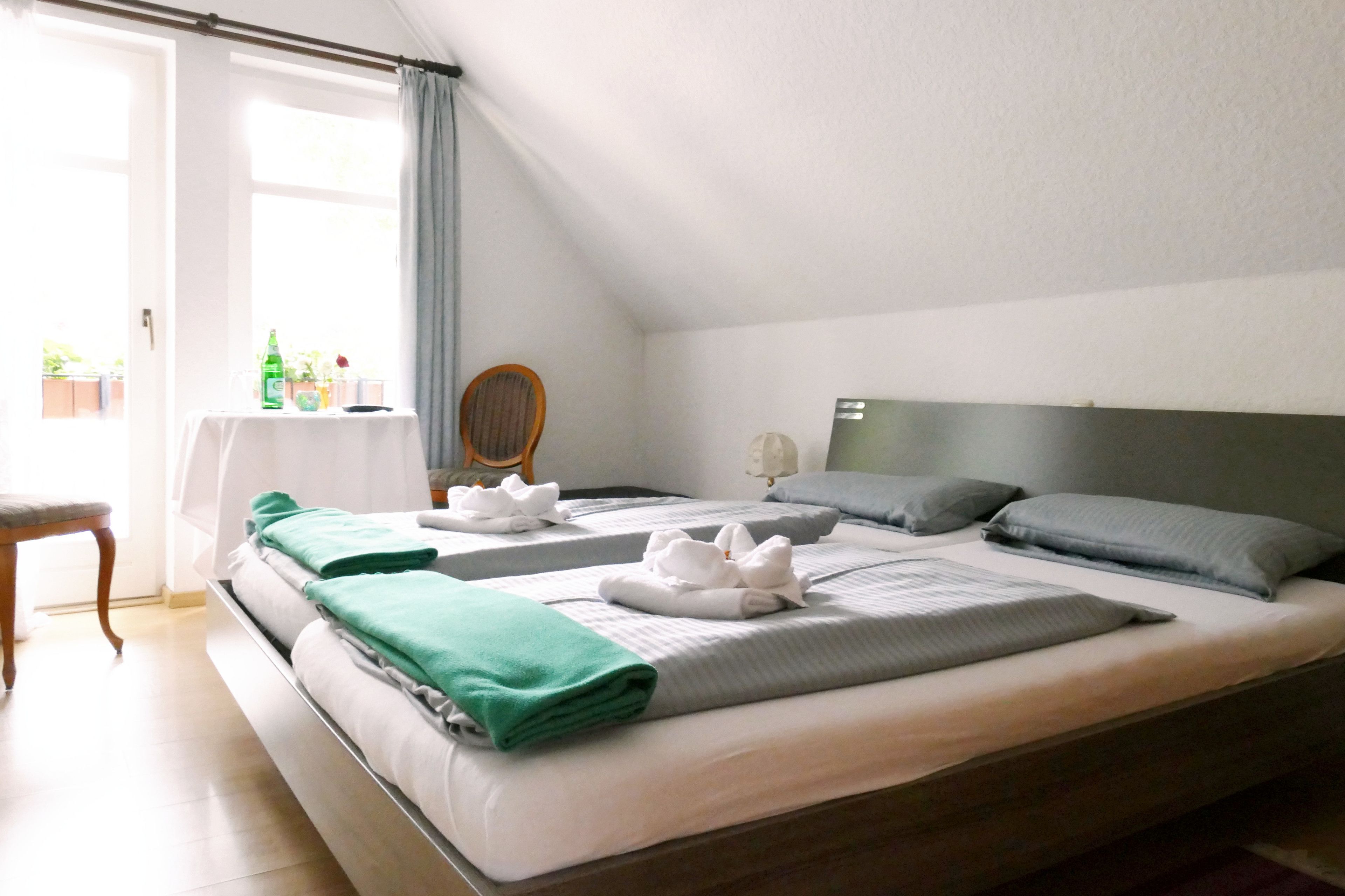 Quarto de hotel com duas camas, uma cadeira, uma mesa com abajur e uma janela com cortinas.