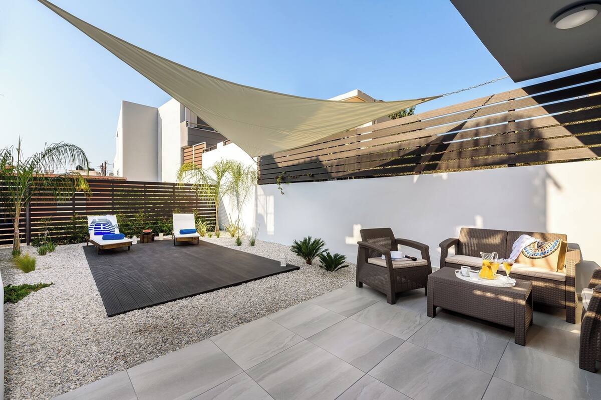 Uma área de estar moderna ao ar livre, com sofá, cadeiras e uma mesa, cercada por um cercado de madeira e plantas.