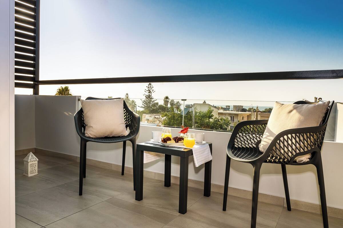 Uma varanda com duas cadeiras, uma mesa pequena com uma bandeja de comida e bebidas, e vista para o horizonte urbano.