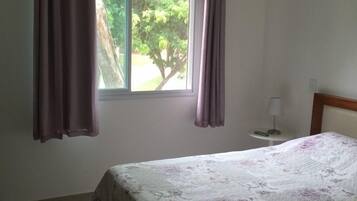 Quarto com cama, janela com cortinas, mesa de cabeceira com abajur e uma cadeira.