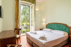 Quarto de hotel com cama, escrivaninha, cadeira, mesa de cabeceira e janela com cortinas.