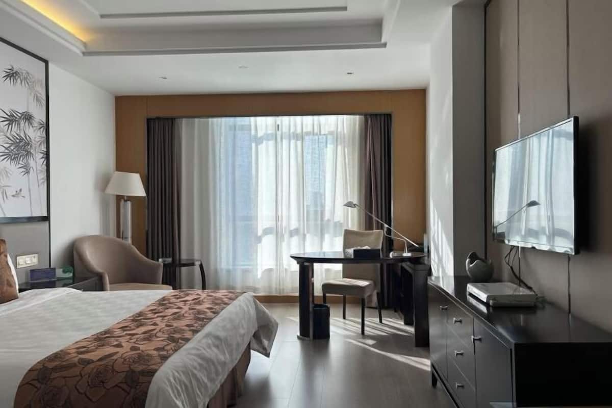 Quarto de hotel com cama, escrivaninha, cadeira, televisão e janelas amplas.