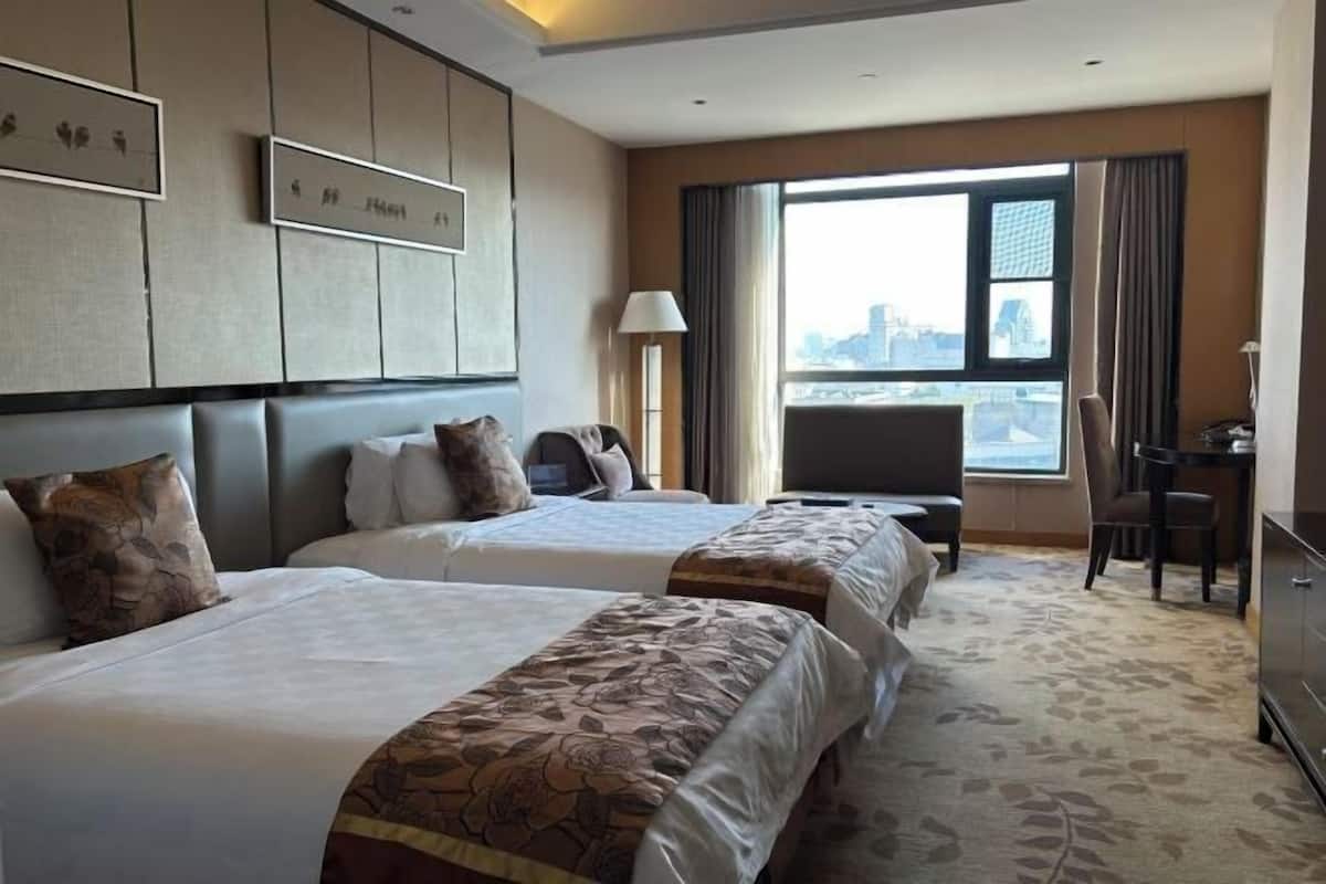 Quarto de hotel com duas camas, uma escrivaninha, uma cadeira, um abajur e uma janela grande com vista para a cidade.