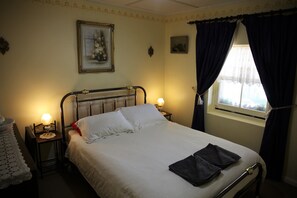 Uma cama bem arrumada com lençóis brancos, duas luminárias de cabeceira, um quadro emoldurado acima da cama e uma janela com cortinas.
