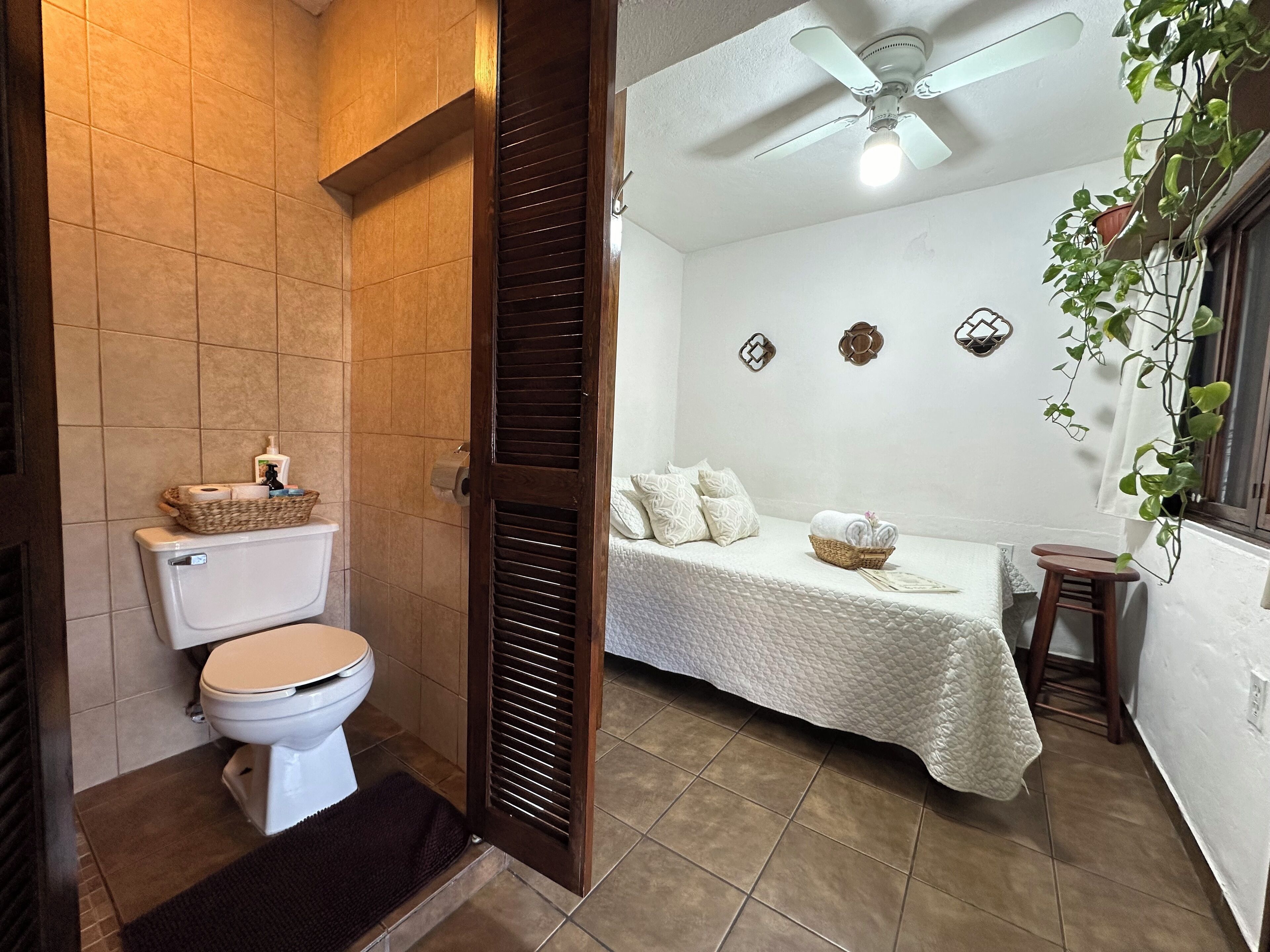 Casa de banho com sanita, zona de duche e cama com colcha branca.