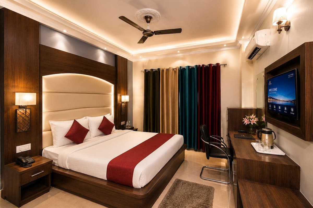 Quarto de hotel com cama, mesa de cabeceira, escrivaninha com cadeira, televisão e ventoinha de teto.