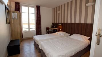 Quarto de hotel com duas camas, uma escrivaninha, uma cadeira e uma janela com cortinas.