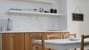 Cozinha com bancada branca, armários de madeira e mesa de jantar com cadeiras. Há prateleiras acima da bancada com diversos utensílios de cozinha.