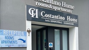 Entrada de um prédio com uma placa indicando 'Constantino Home rooms & apartments' e uma placa de 'Reception Rooms Apartments'.